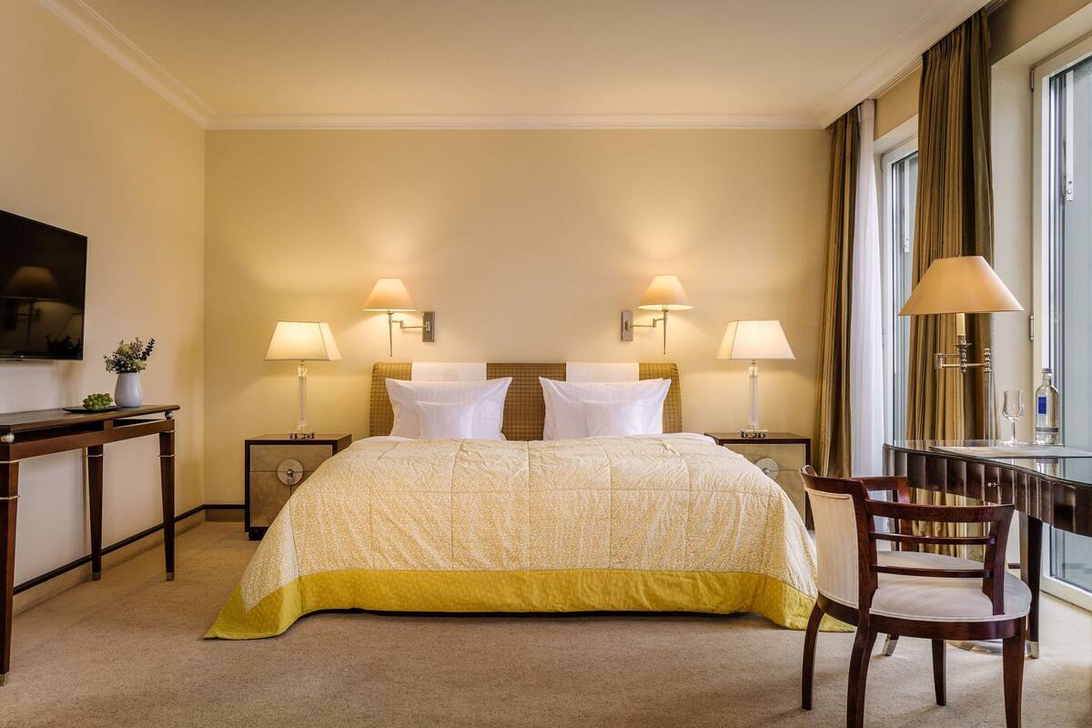 Quarto de hotel com uma cama grande, duas mesas de cabeceira, uma escrivaninha com cadeira e uma TV na parede.