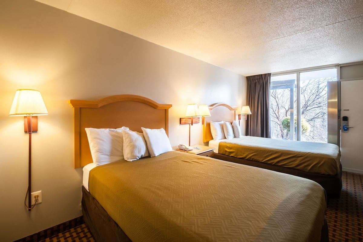 Quarto de hotel com duas camas, cada uma com cabeceira e luminárias laterais. Há uma janela com cortinas e uma porta com puxador.