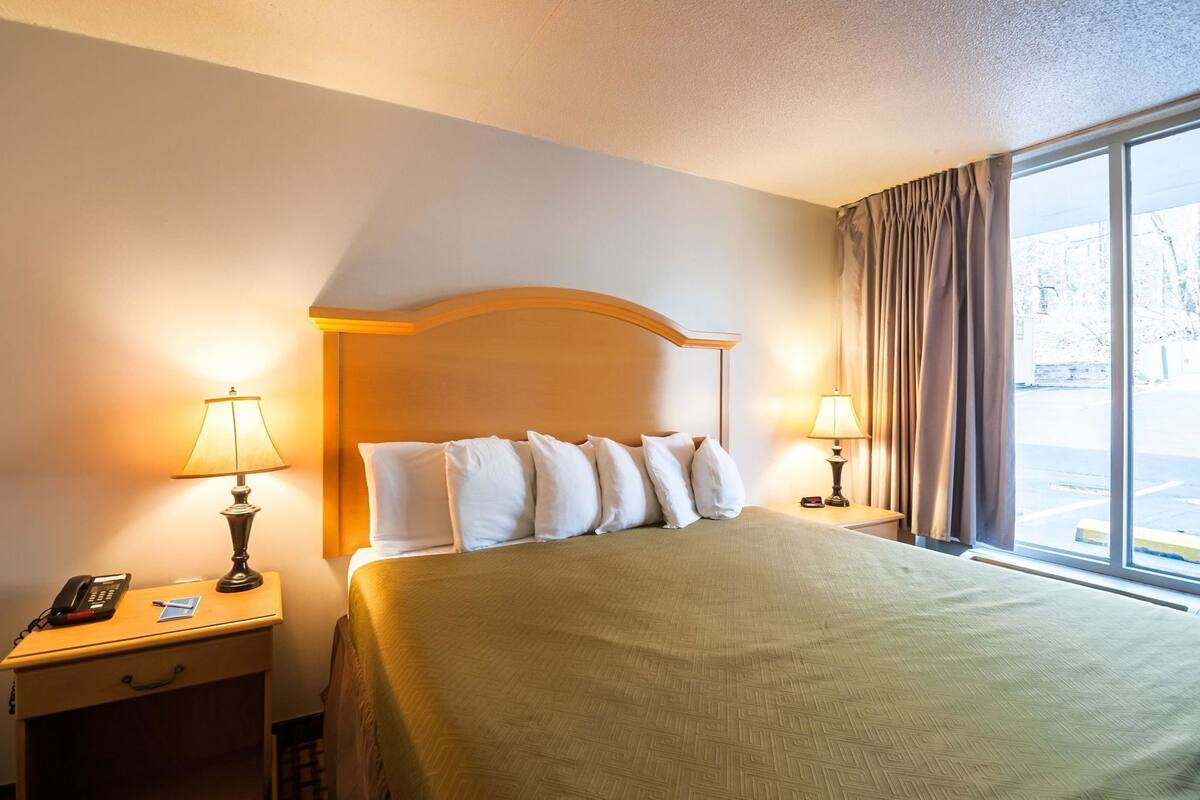 Quarto de hotel com cama, duas luminárias de cabeceira, mesa de apoio com telefone e janela com cortinas.