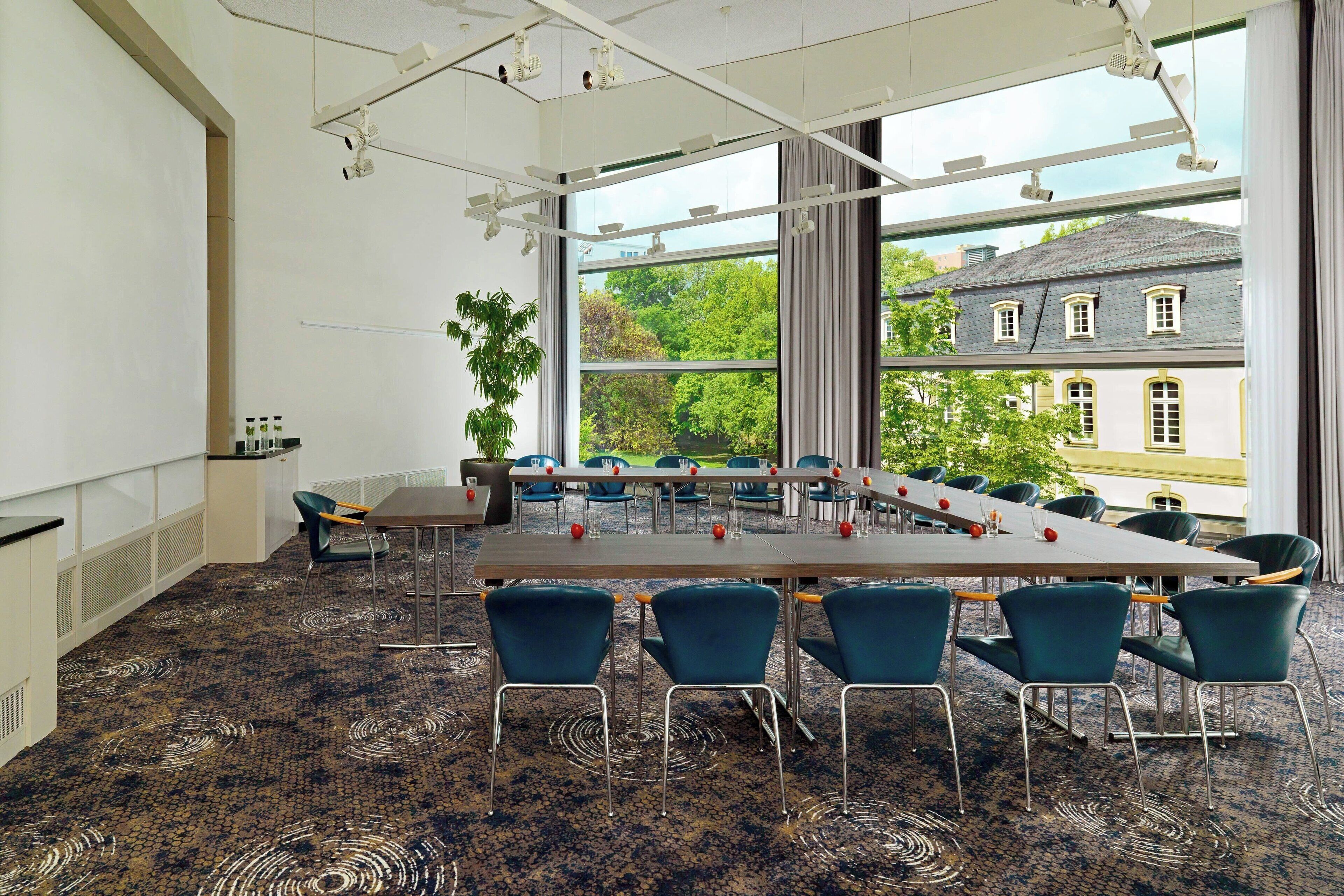 Sala de conferência com uma mesa grande, cadeiras azuis e vista para um edifício através de janelas amplas.