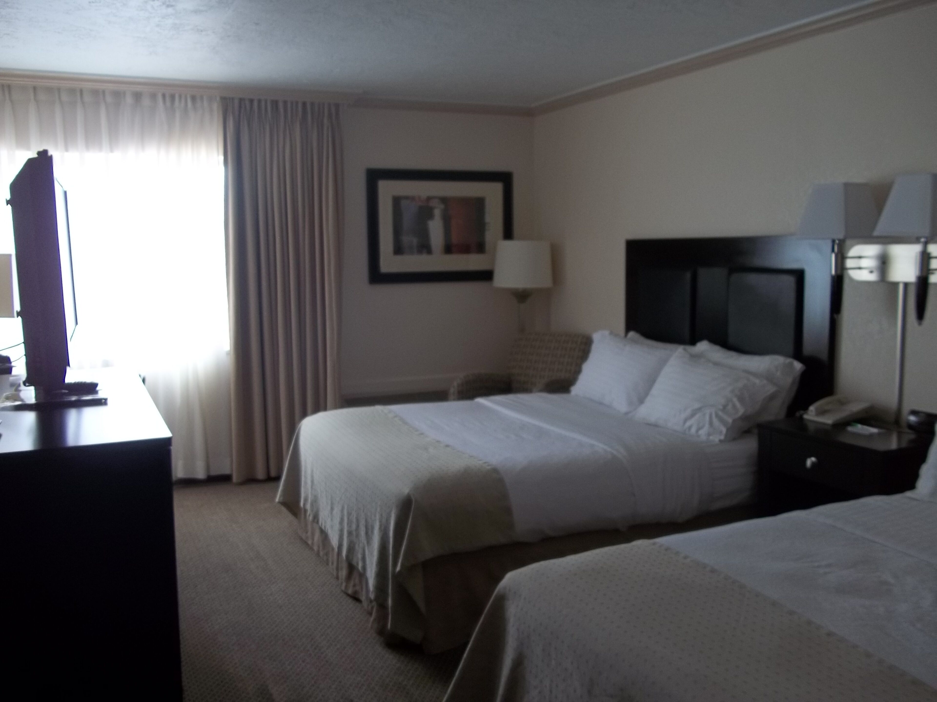 Quarto de hotel com duas camas, uma escrivaninha com computador, uma cadeira, um abajur e um quadro na parede.