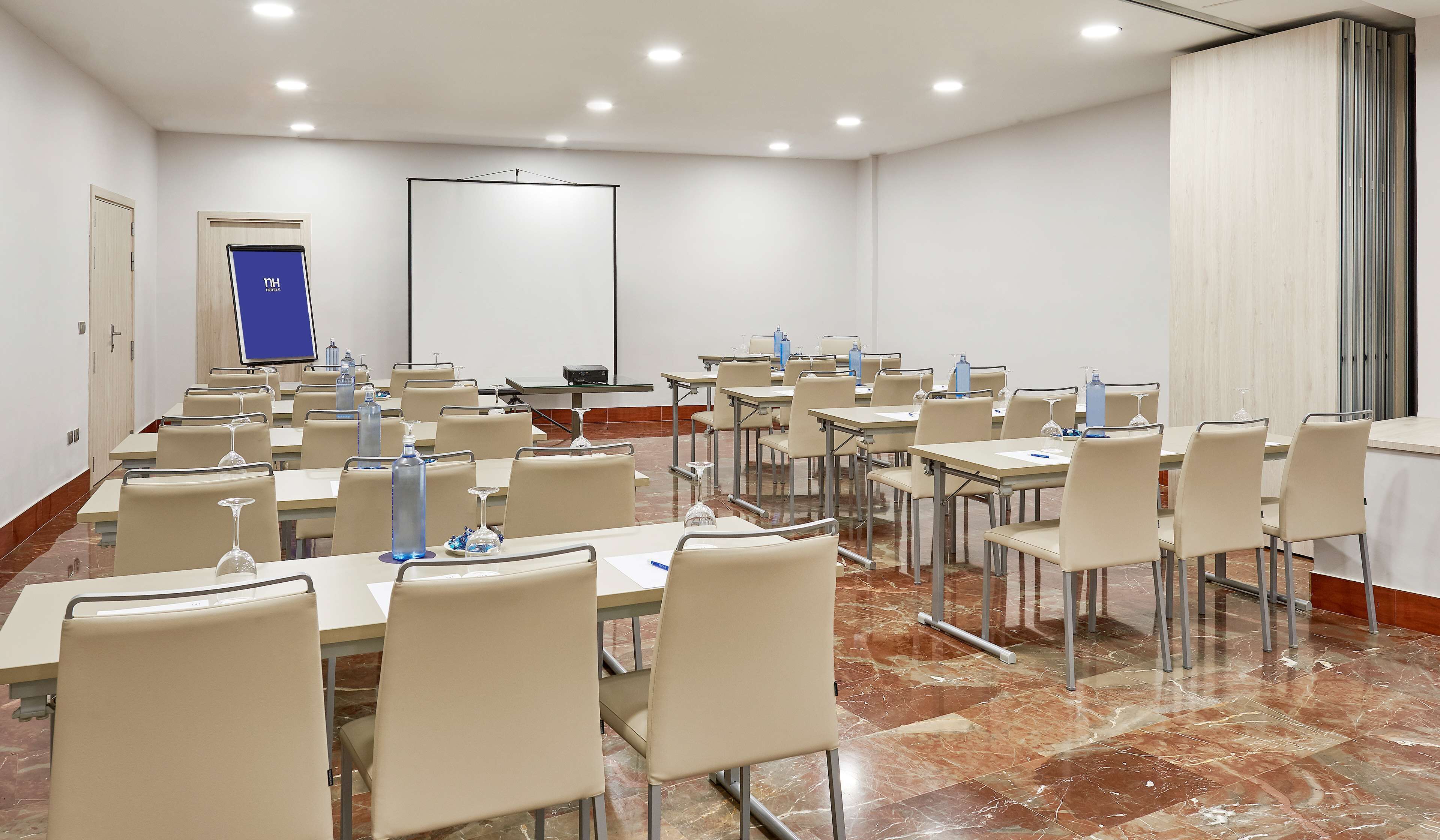 Sala de conferência com várias mesas e cadeiras, equipada com garrafas de água e copos, e uma tela de projeção.