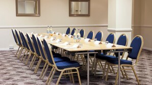 Sala de conferência com uma mesa comprida, cadeiras azuis, cartões de identificação, garrafas de água e piso coberto por carpete.
