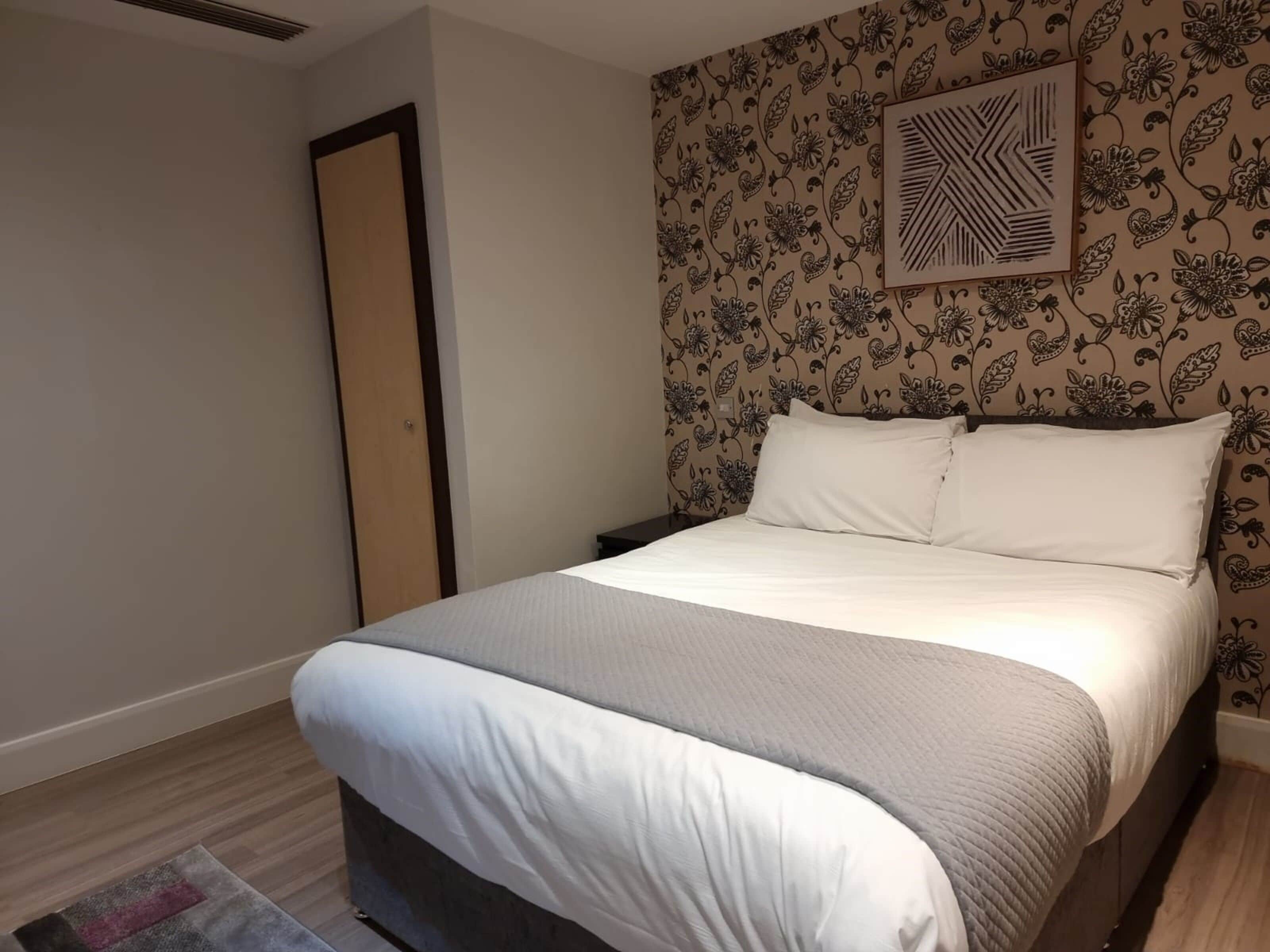 Cama bem arrumada com lençóis brancos e manta cinza em quarto com papel de parede floral e uma obra de arte geométrica na parede.