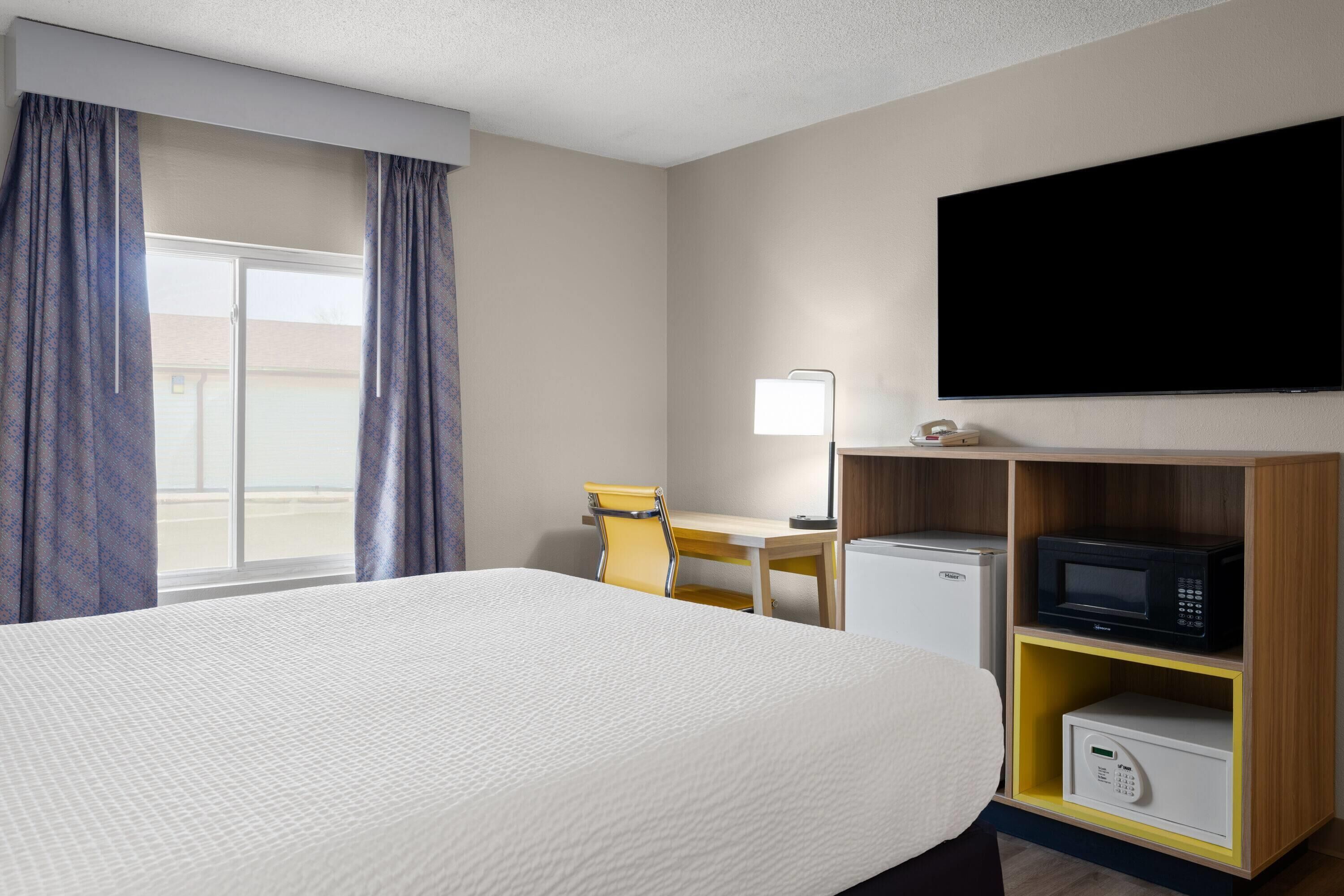 Quarto de hotel com cama, escrivaninha, cadeira, televisão, micro-ondas e mini-geladeira.