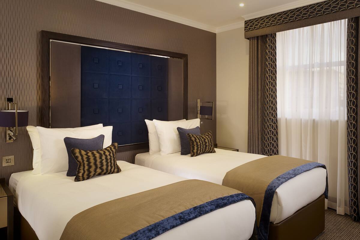 Quarto de hotel com duas camas, uma janela grande com cortinas e uma parede com padrão geométrico.