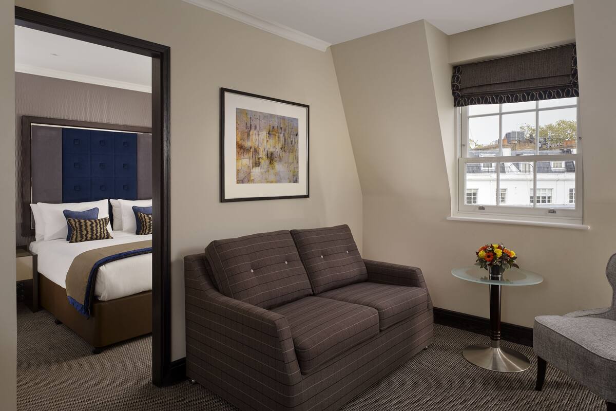 Quarto de hotel com cama, um sofá marrom, uma poltrona cinza, uma pequena mesa com flores e uma janela com vista para edifícios.