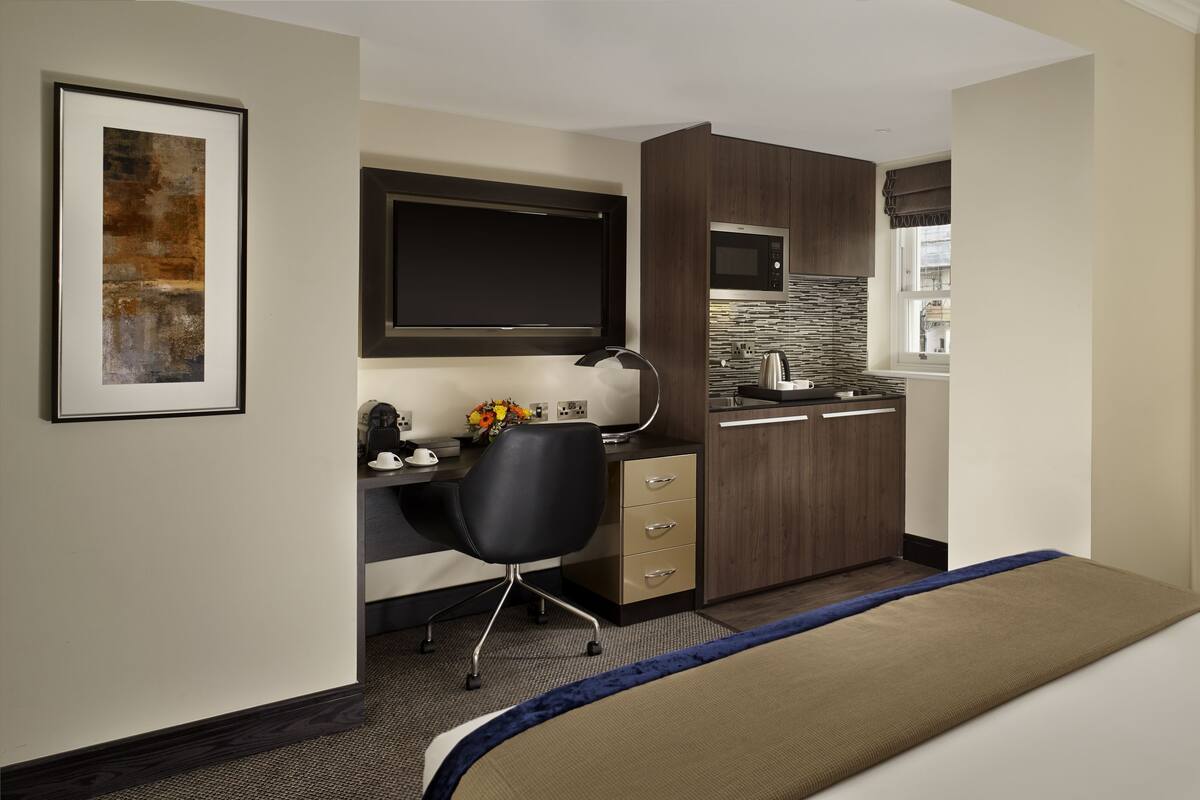 Quarto de hotel com cama, escrivaninha, cadeira, micro-ondas, pia e janela.
