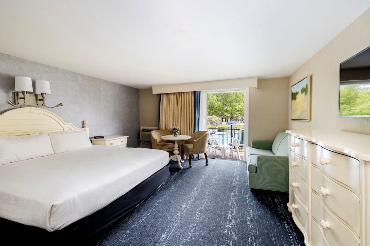 Quarto de hotel com uma cama grande, uma mesinha com cadeiras, uma poltrona verde, um armário e uma varanda com vista para árvores.