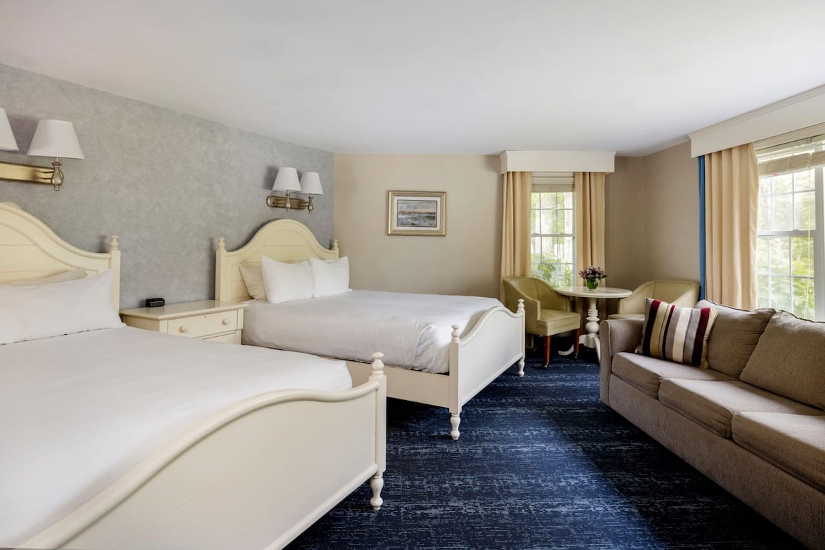 Quarto de hotel com duas camas, um sofá, uma escrivaninha e uma janela com cortinas.
