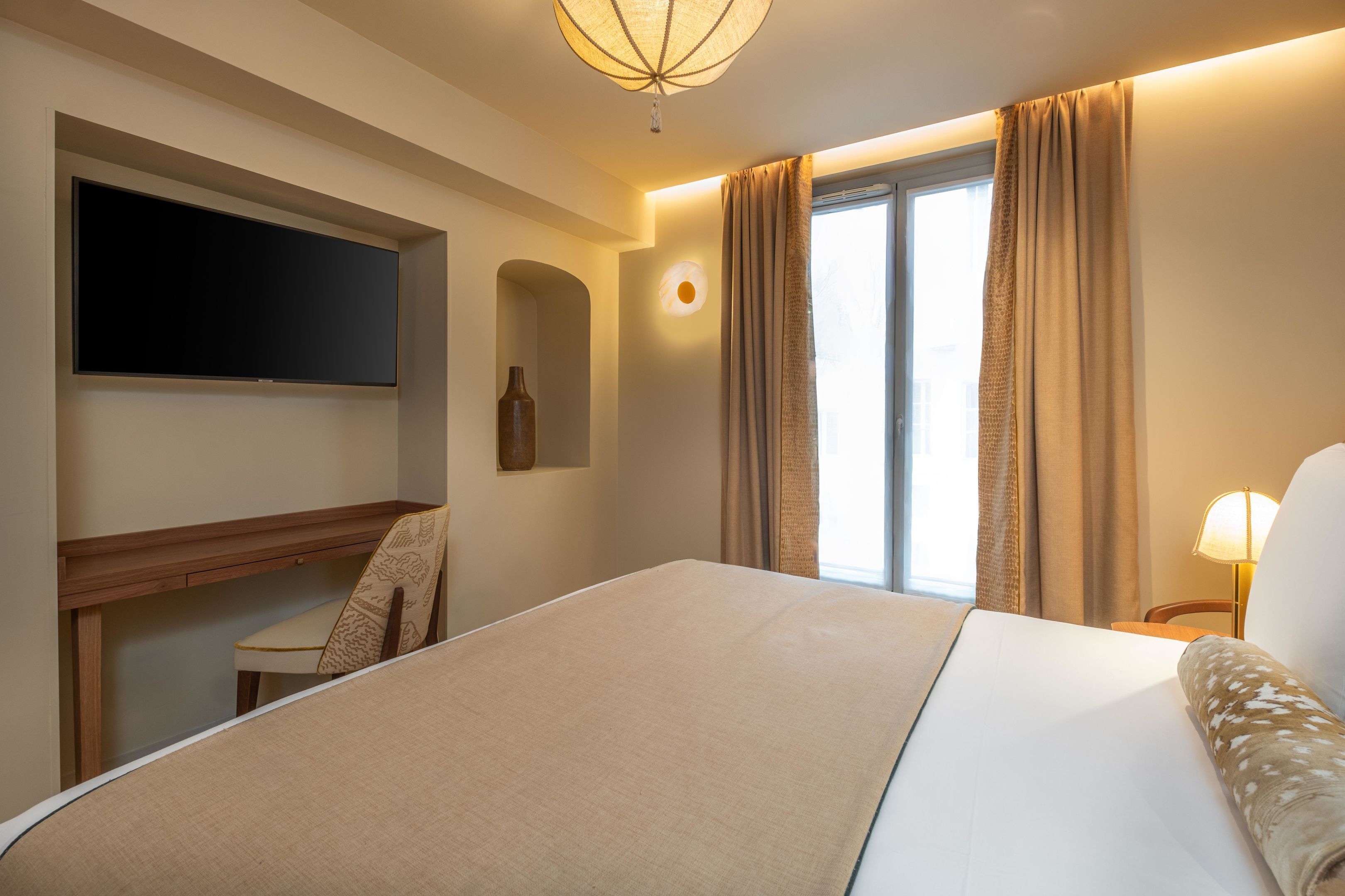 Quarto de hotel com uma cama grande, televisão de tela plana na parede, mesa de cabeceira com abajur e uma janela com cortinas.