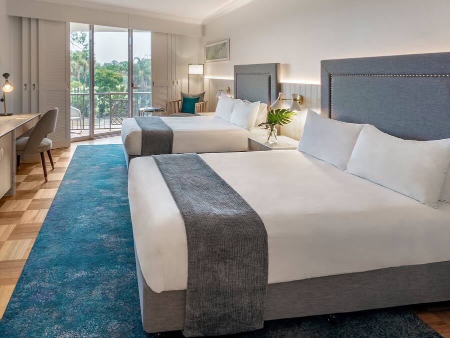 Quarto de hotel com duas camas, uma escrivaninha com televisão, uma cadeira, varanda com vista e uma janela grande.