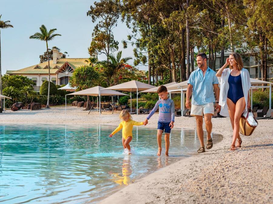 Uma família caminhando pela praia, com uma piscina, palmeiras e um edifício de resort ao fundo.