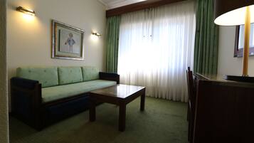 Quarto com sofá verde, mesa de centro de madeira e uma luminária. Há um quadro na parede e uma janela com cortinas verdes.