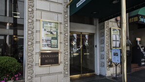 Exterior do Chancellor Hotel, com entrada de vidro, uma placa de identificação e um caixa eletrônico à direita.