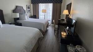 Quarto de hotel com duas camas, uma escrivaninha, uma cadeira, um ventilador de teto e uma janela com cortinas.