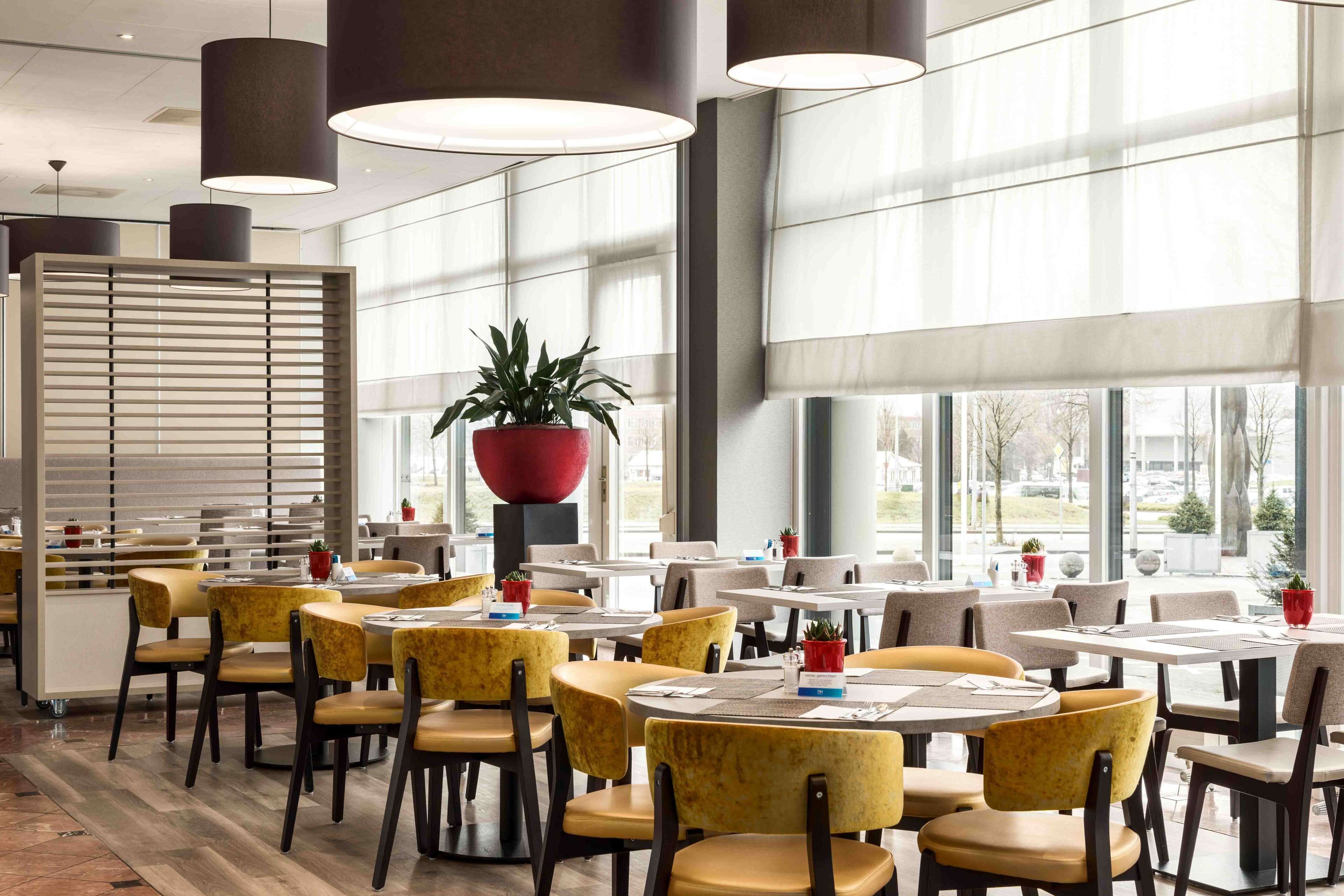 Um restaurante moderno com mesas redondas e cadeiras amarelas, janelas amplas e plantas em vasos.