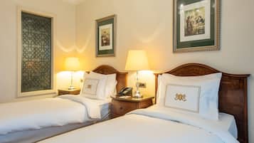 Quarto de hotel com duas camas, cada uma com roupa de cama branca e almofadas decorativas, cabeceiras de madeira, mesas de cabeceira com abajures e quadros nas paredes.