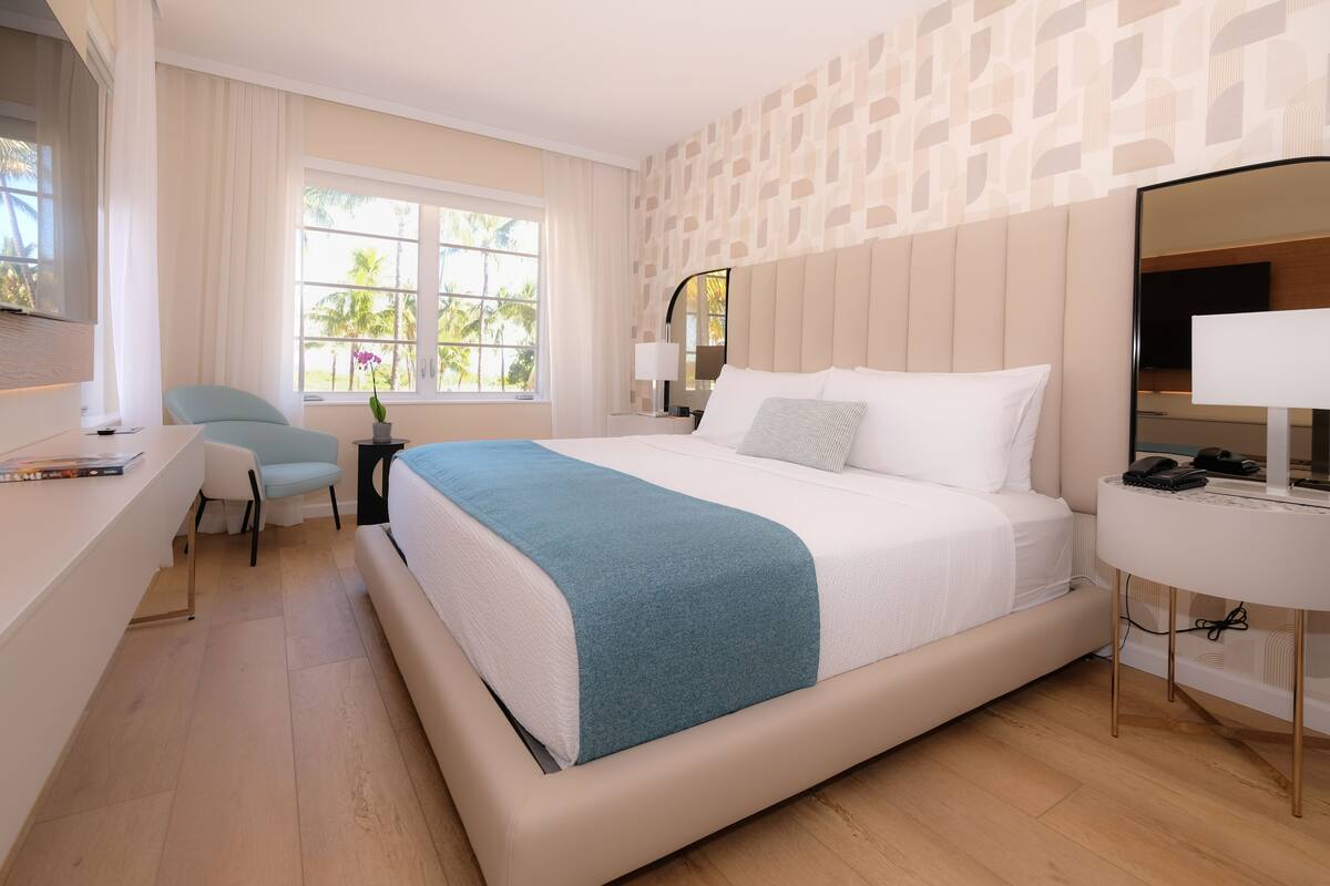 Quarto de hotel moderno com uma cama grande, uma escrivaninha, uma cadeira, uma mesa de cabeceira com luminária e uma janela ampla com vista para palmeiras.