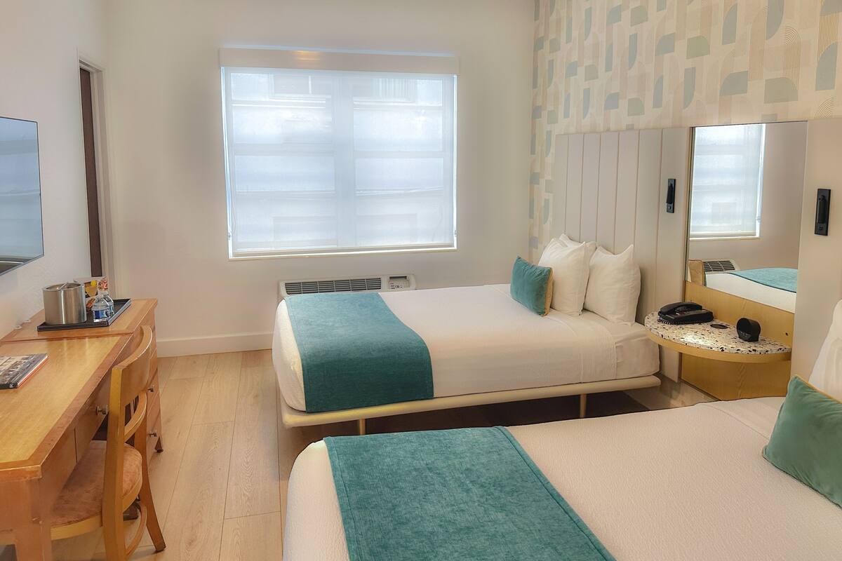 Quarto de hotel com cama, escrivaninha, cadeira, espelho e janela.