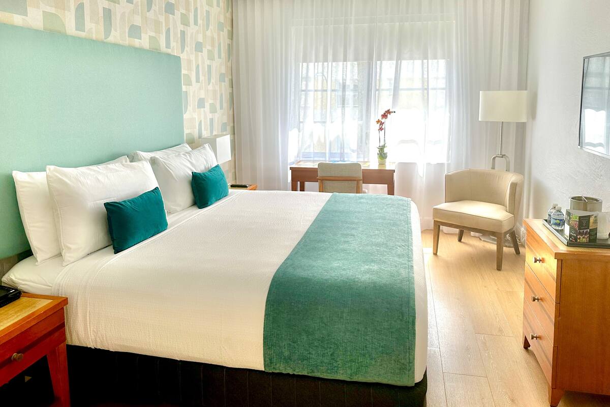 Quarto de hotel com uma cama grande, uma escrivaninha com cadeira, uma luminária e um armário.