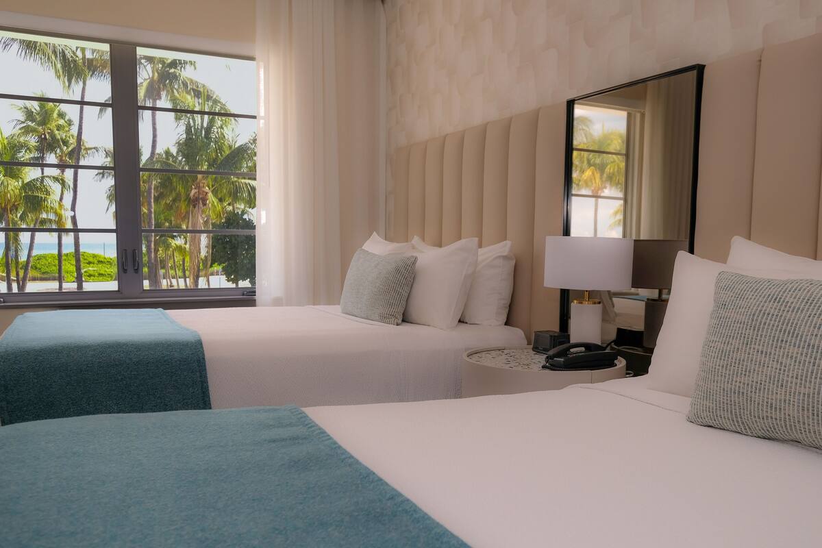 Quarto de hotel com duas camas, uma janela ampla com vista para palmeiras e o mar, mesa de cabeceira com luminária e espelho na parede.