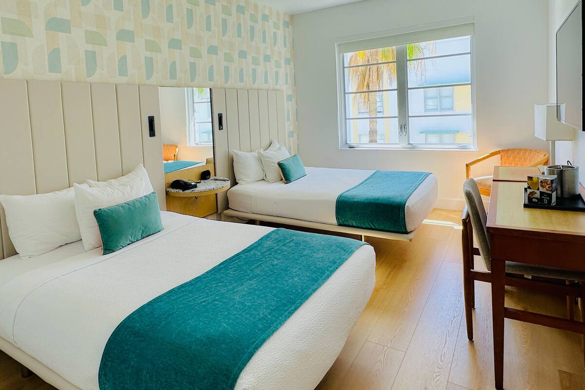 Quarto de hotel com duas camas, uma escrivaninha, uma cadeira, mesa de cabeceira, abajur e janela com vista para palmeiras.