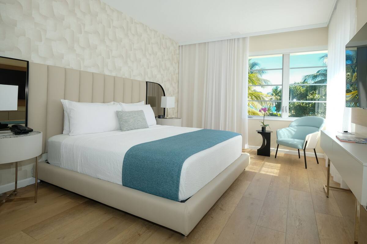 Quarto de hotel com uma cama grande, uma escrivaninha, uma cadeira e vista para palmeiras pela janela.
