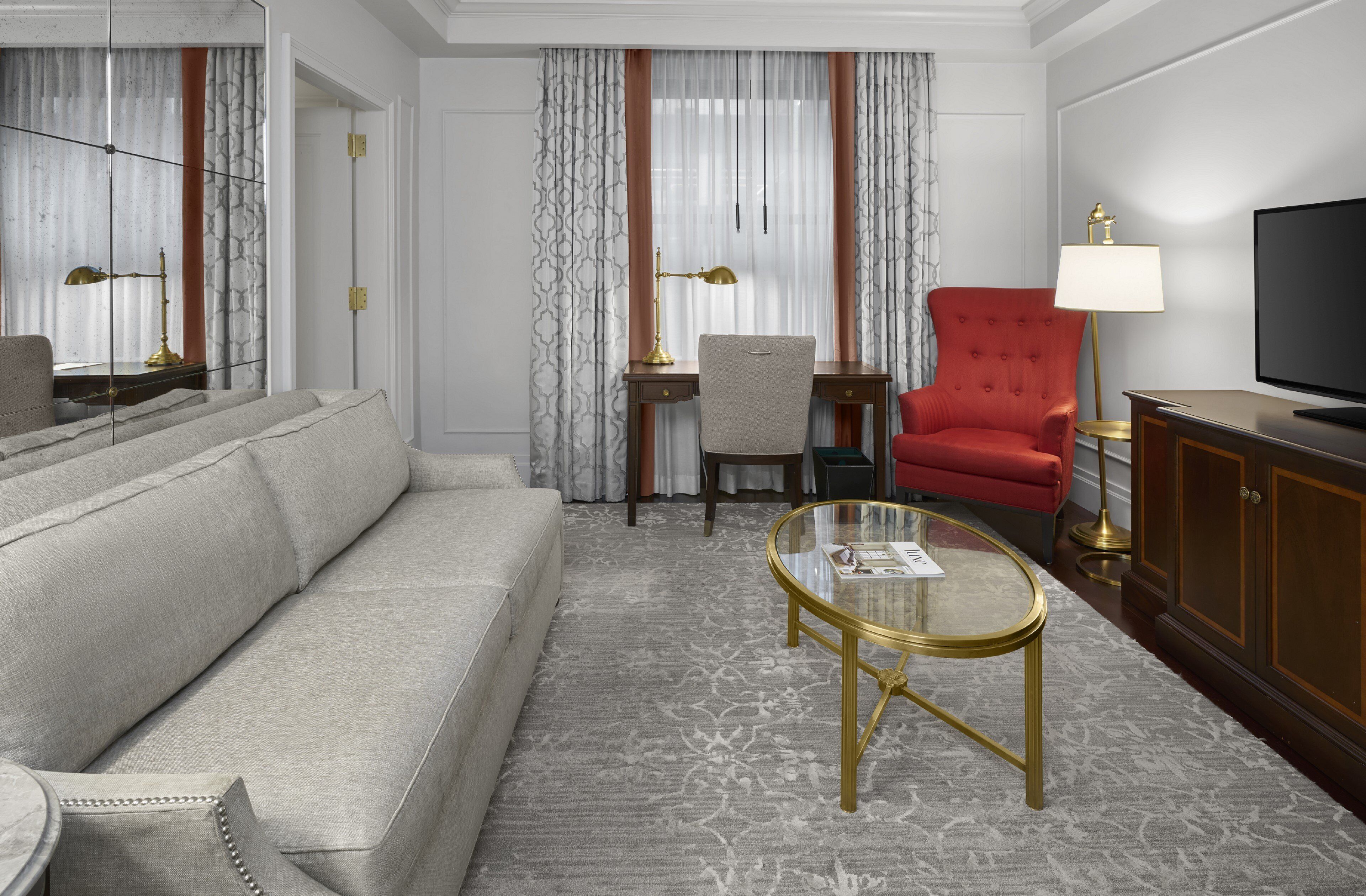 Quarto de hotel moderno com sofá, mesa de centro de vidro, poltrona vermelha, televisão e escrivaninha com abajur.