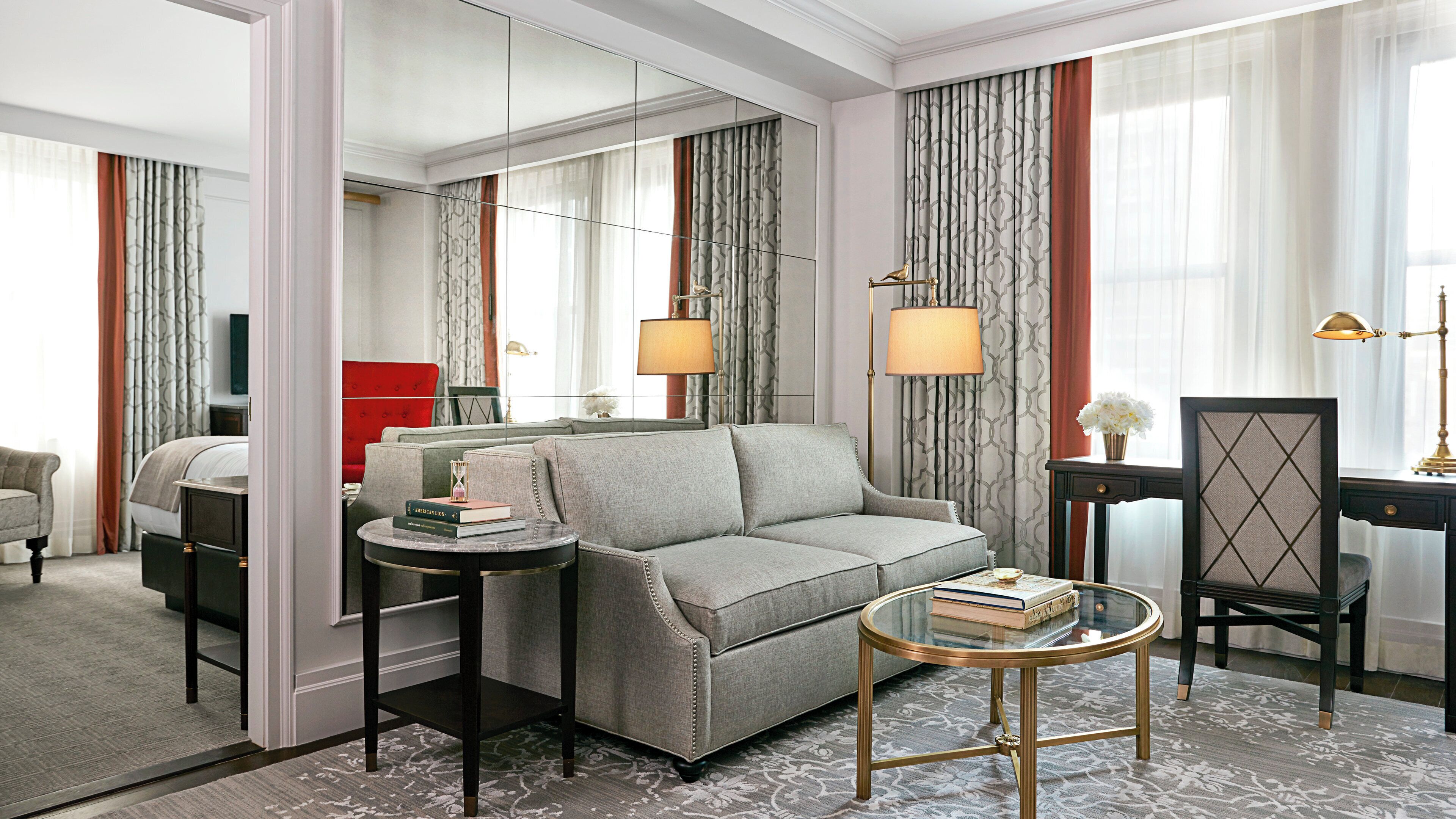Quarto de hotel moderno com um sofá cinzento, uma mesa de centro redonda de vidro e uma poltrona vermelha.