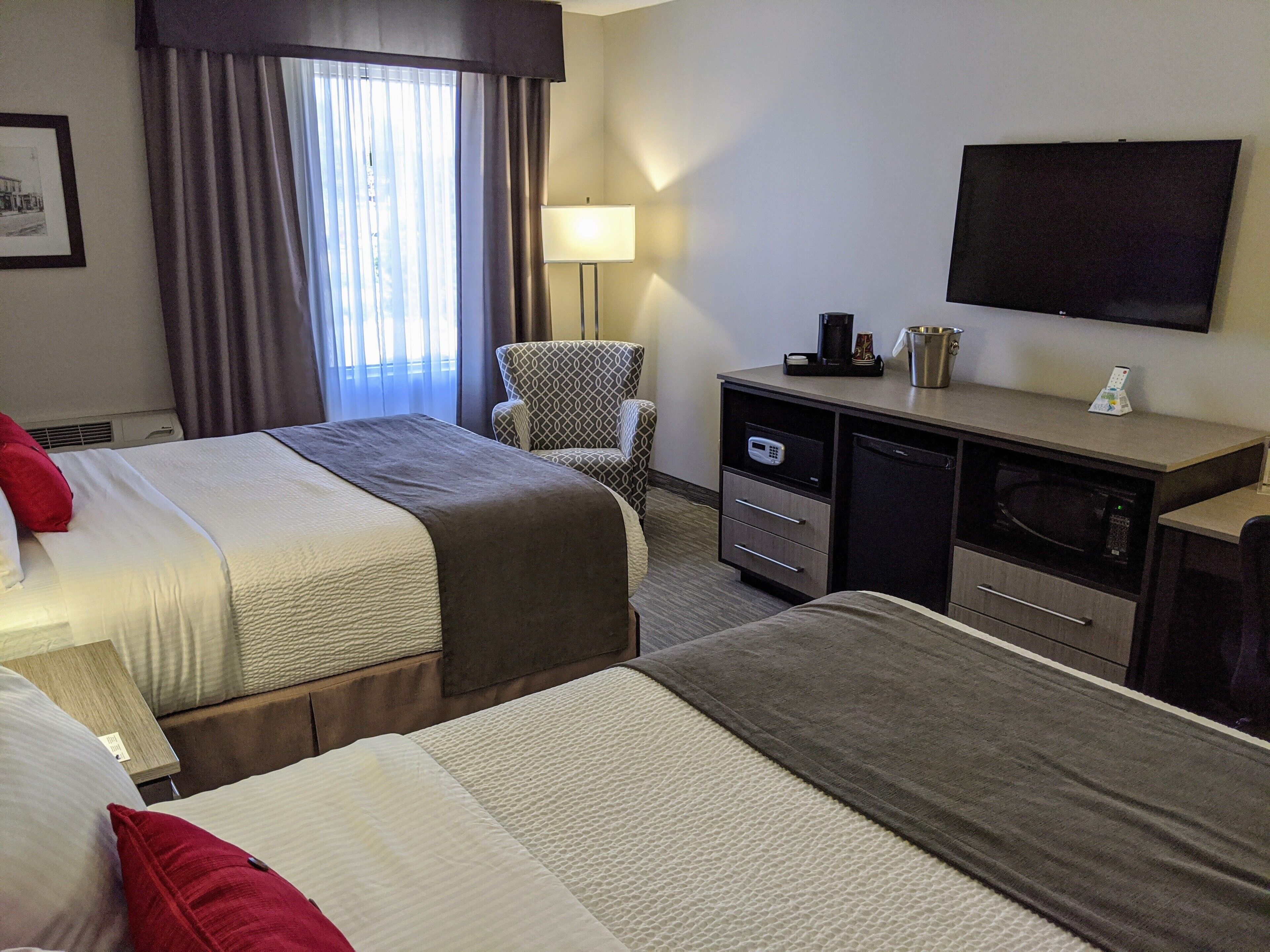 Quarto de hotel com duas camas, uma escrivaninha com micro-ondas, uma cadeira, televisão e janela com cortinas.