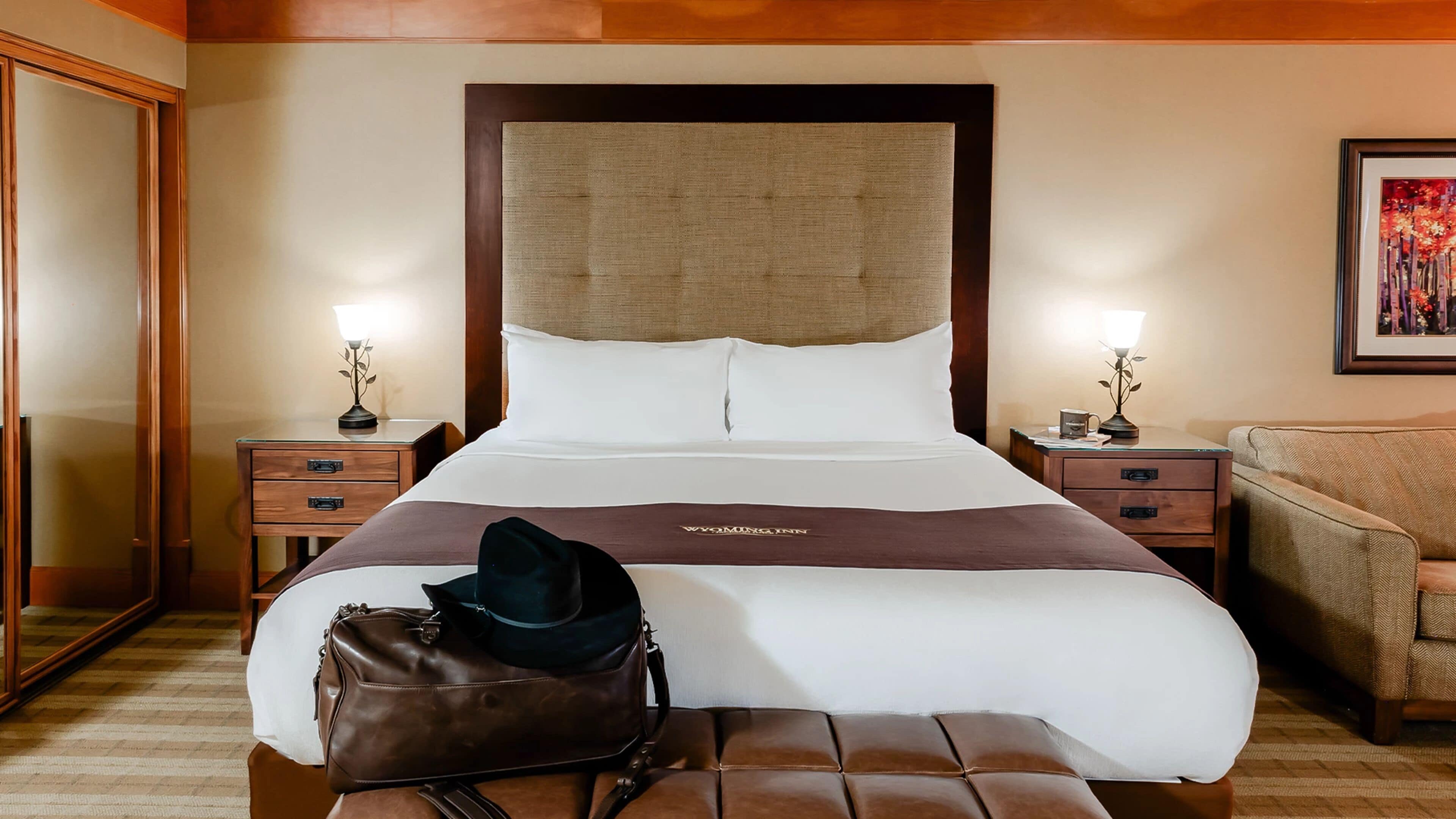 Quarto de hotel com uma cama grande, duas mesas de cabeceira com abajures, uma mala de couro marrom na cama e um banquinho marrom.