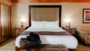 Quarto de hotel com uma cama grande, duas mesas de cabeceira com abajures, uma mala de couro marrom na cama e um banquinho marrom.
