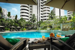 Área da piscina com espreguiçadeiras, uma mesa com uma cesta de frutas e bebidas, e vista para edifícios altos ao fundo.