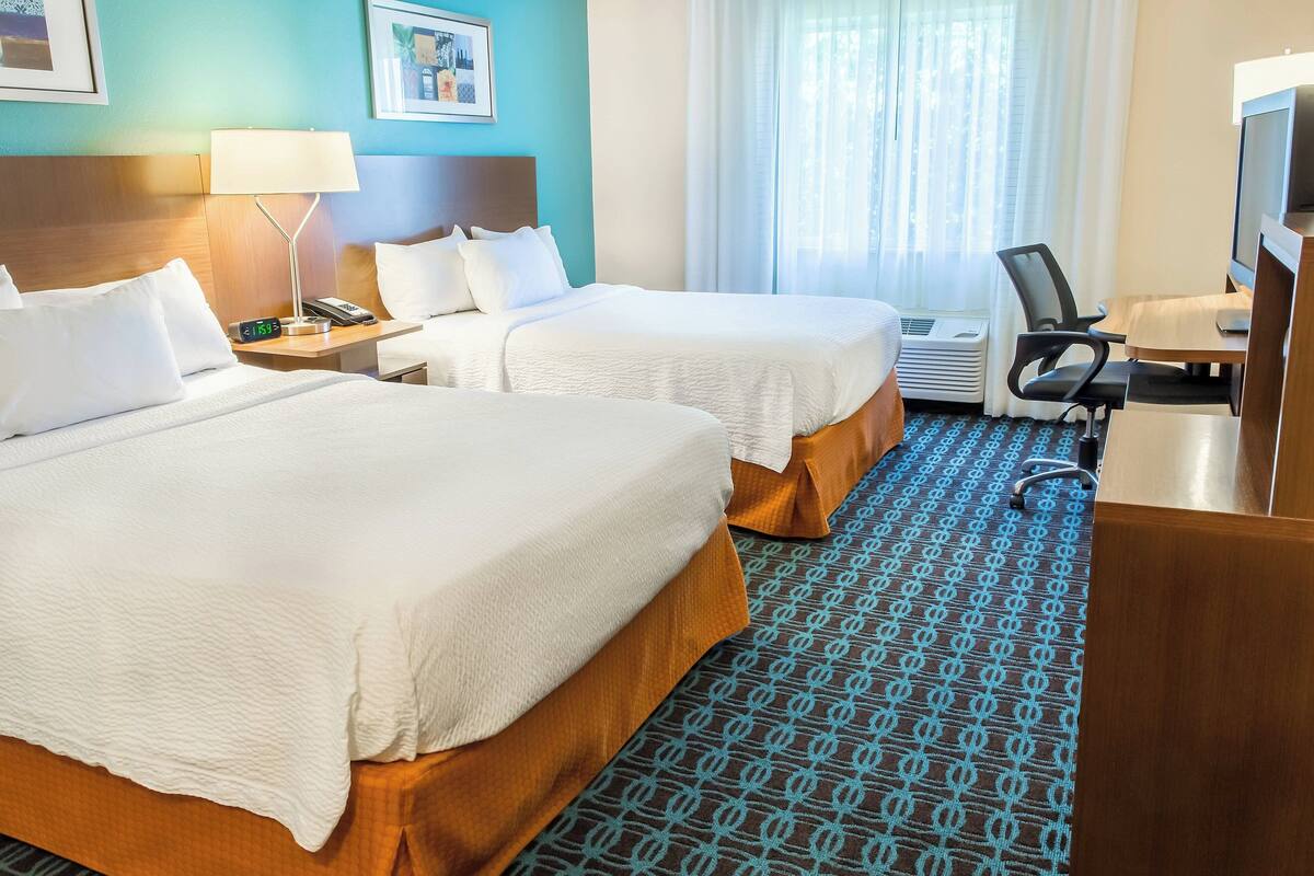 Quarto de hotel com duas camas, uma mesa de escritório com um computador, uma luminária e uma janela com cortinas.