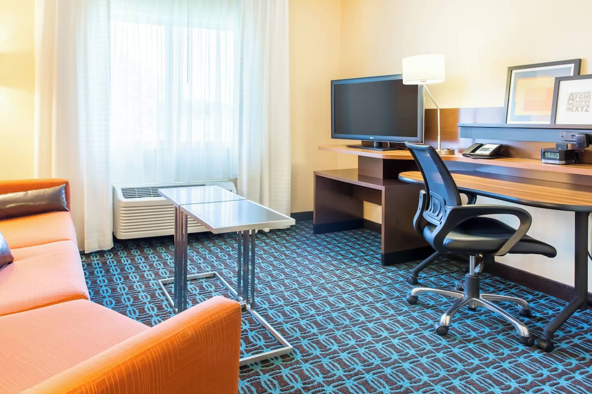 Quarto de hotel com cama, escrivaninha com computador e TV, uma cadeira e janela com cortinas.