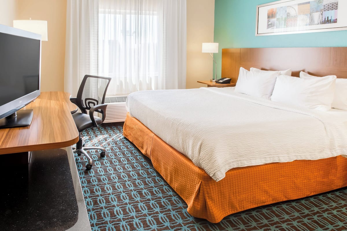 Quarto de hotel com uma cama, uma escrivaninha com televisão, uma cadeira e um tapete estampado.