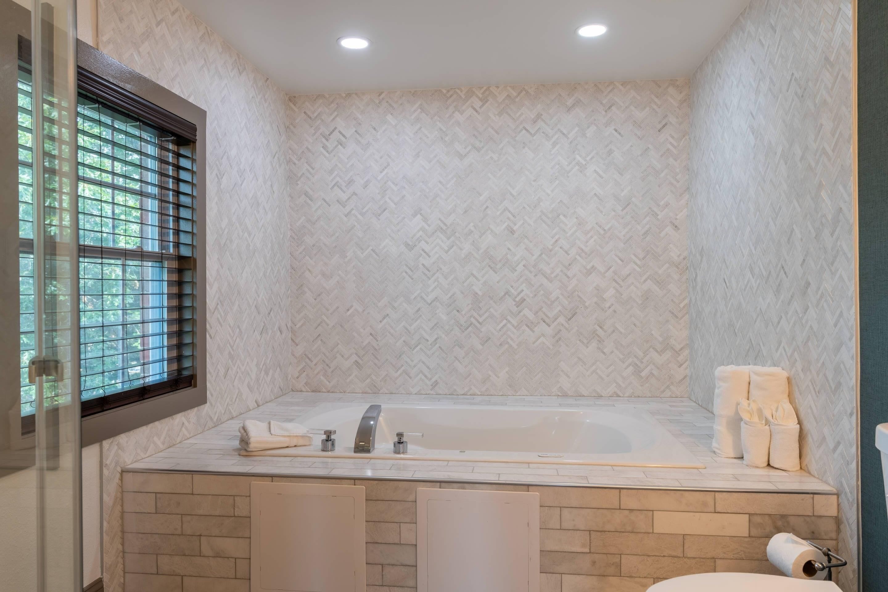 Uma casa de banho com uma banheira grande, uma janela com persianas e uma parede revestida a azulejos.