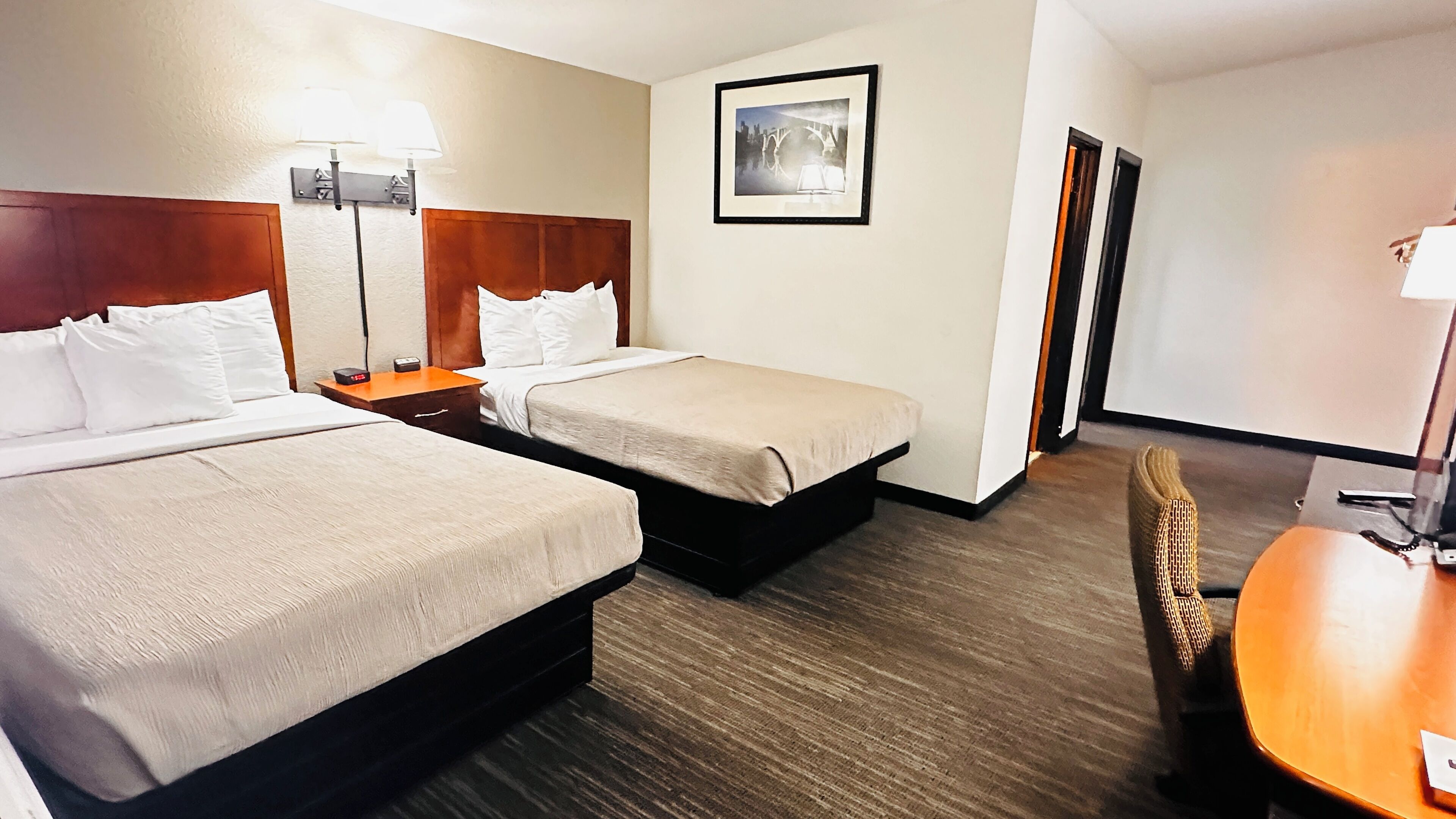 Quarto de hotel com duas camas, uma escrivaninha, uma cadeira, um abajur e um quadro na parede.