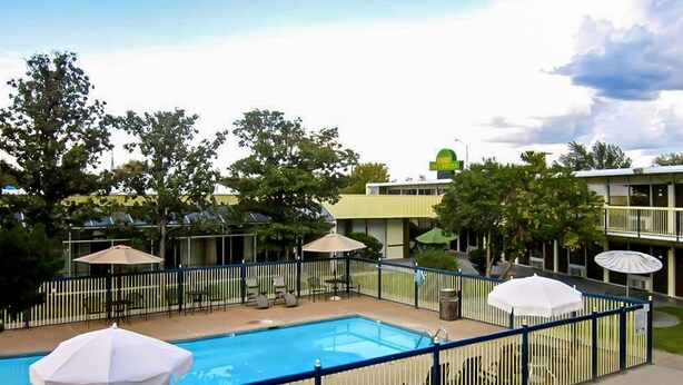 Piscina com espreguiçadeiras e guarda-sóis, cercada por uma grade, com um edifício ao fundo.