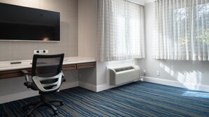 Quarto de hotel com televisão de tela plana na parede, uma escrivaninha com cadeira, janela com cortinas e um tapete azul.