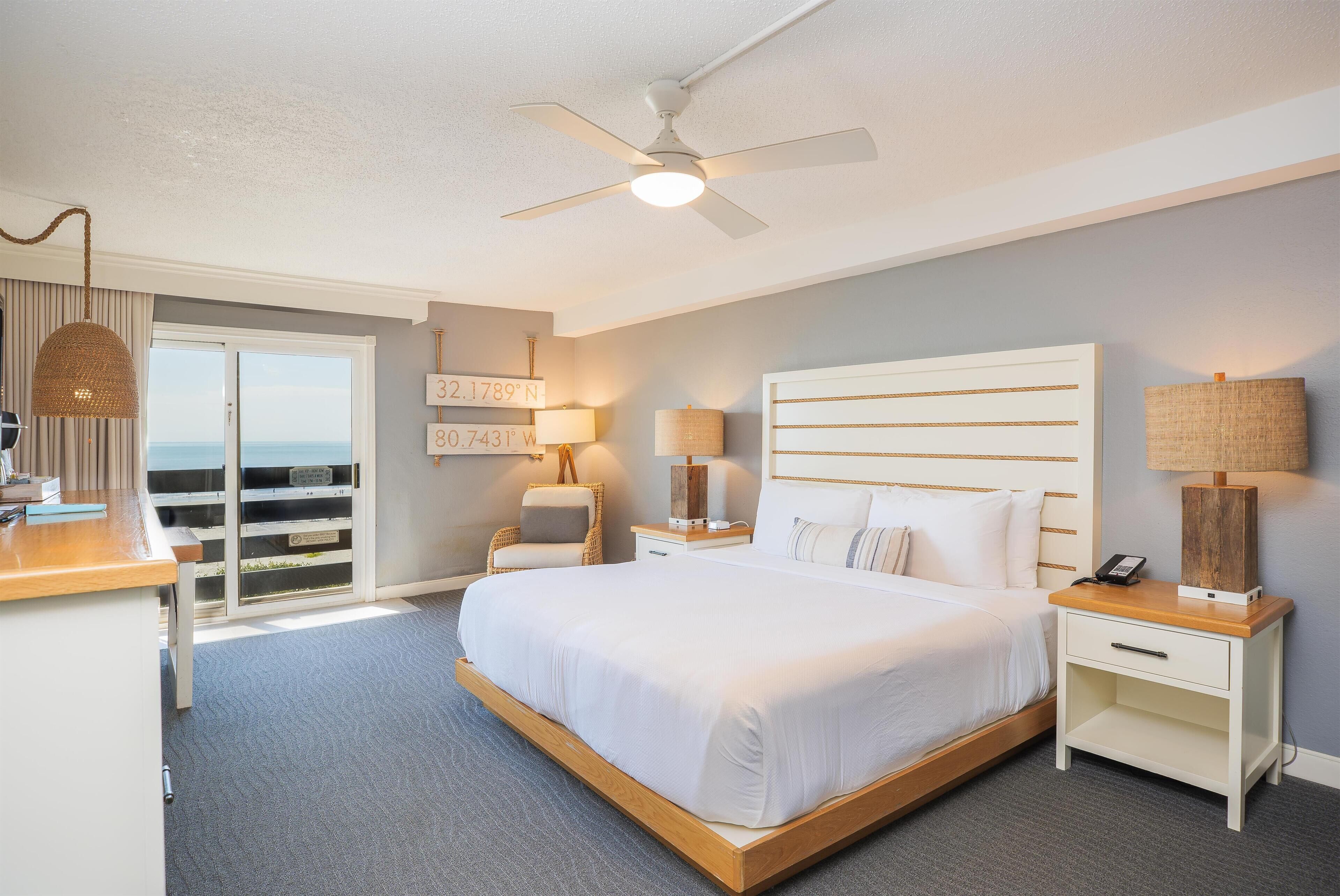 Quarto de hotel com cama, mesas de cabeceira, escrivaninha, cadeira e ventoinha no teto, além de vista para o oceano.
