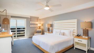 Quarto de hotel com cama, mesas de cabeceira, escrivaninha, cadeira e ventoinha no teto, além de vista para o oceano.
