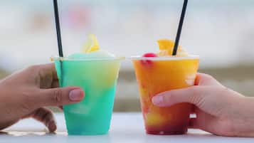 Duas taças de plástico com canudos, uma com uma bebida azul e outra com uma bebida laranja, sendo seguradas por mãos.