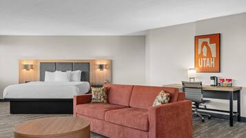 Quarto de hotel com cama, sofá vermelho, mesa de madeira redonda, escrivaninha com cadeira e uma obra de arte na parede.