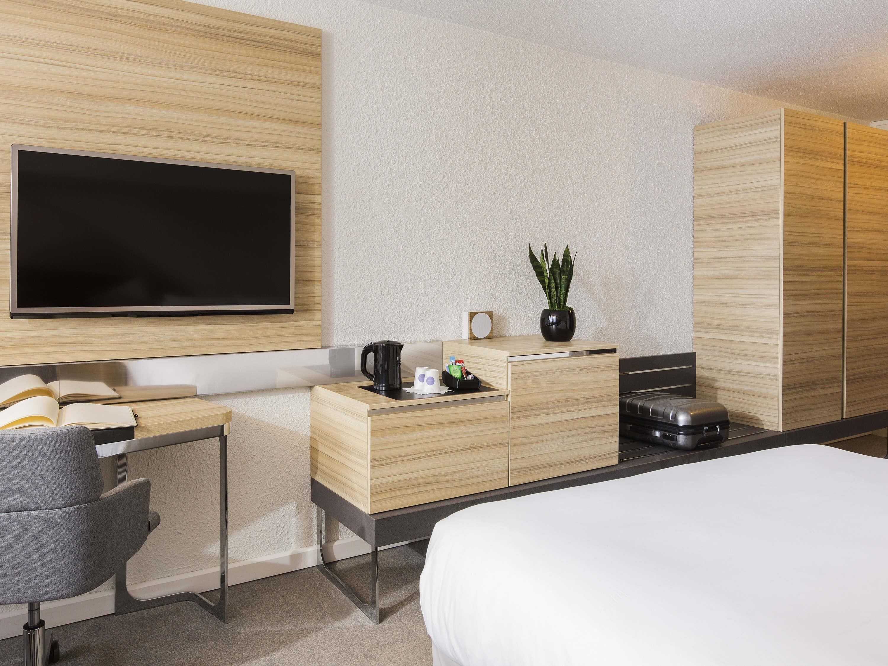 Quarto de hotel moderno com televisão de tela plana, escrivaninha de madeira com máquina de café, planta em vaso e uma cama.