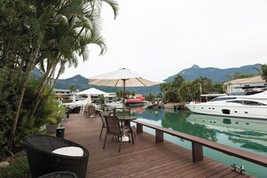 Um deck de madeira com área de estar externa, um areal com barcos e montanhas ao fundo.