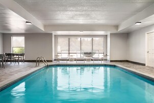 Piscina interior com água azul cristalina, rodeada por espreguiçadeiras e mesas, com amplas janelas que proporcionam iluminação natural.