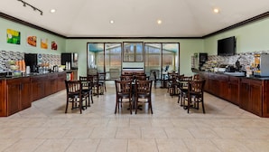 Uma ampla área de refeições com várias mesas e cadeiras, bancada de cozinha com eletrodomésticos e uma grande janela com vista para o exterior.