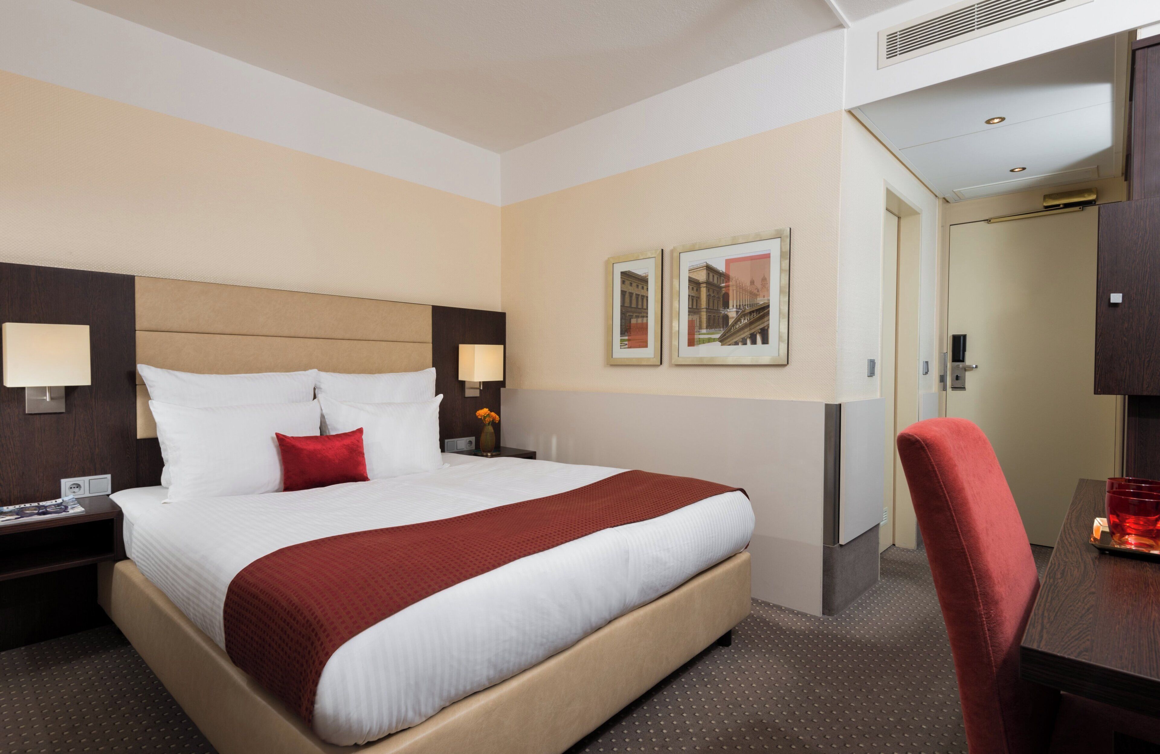 Quarto de hotel com uma cama grande, duas mesas de cabeceira com luminárias, uma escrivaninha com cadeira e quadros nas paredes.